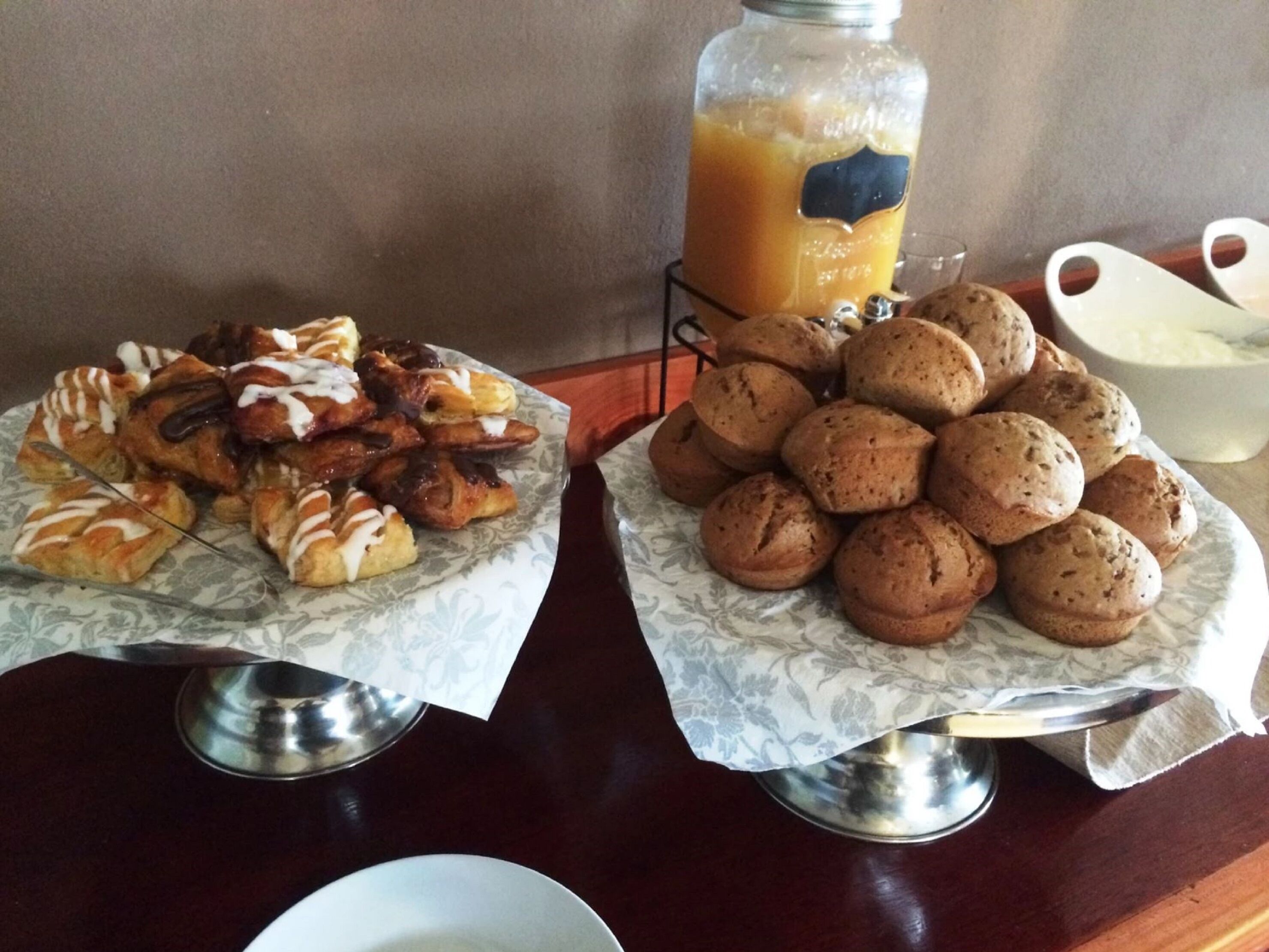 Um buffet de café da manhã com pastelaria e muffins em bandejas de mármore, um pote de vidro com sumo de laranja e uma tigela branca.