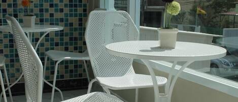 Varanda com cadeiras e mesas de metal branco, parede revestida de azulejos e janela com vista para a rua.