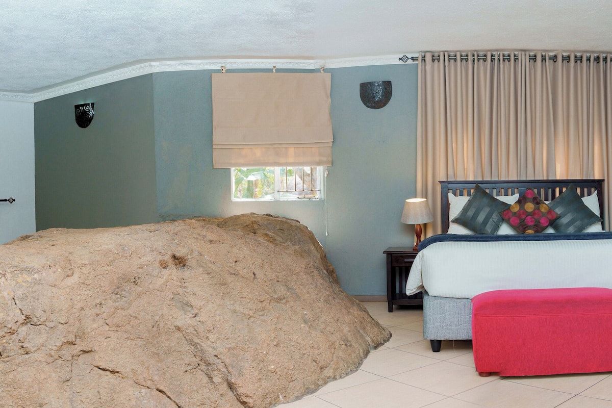 Quarto com uma grande rocha, cama com almofadas, mesa de cabeceira e janela com cortinas.