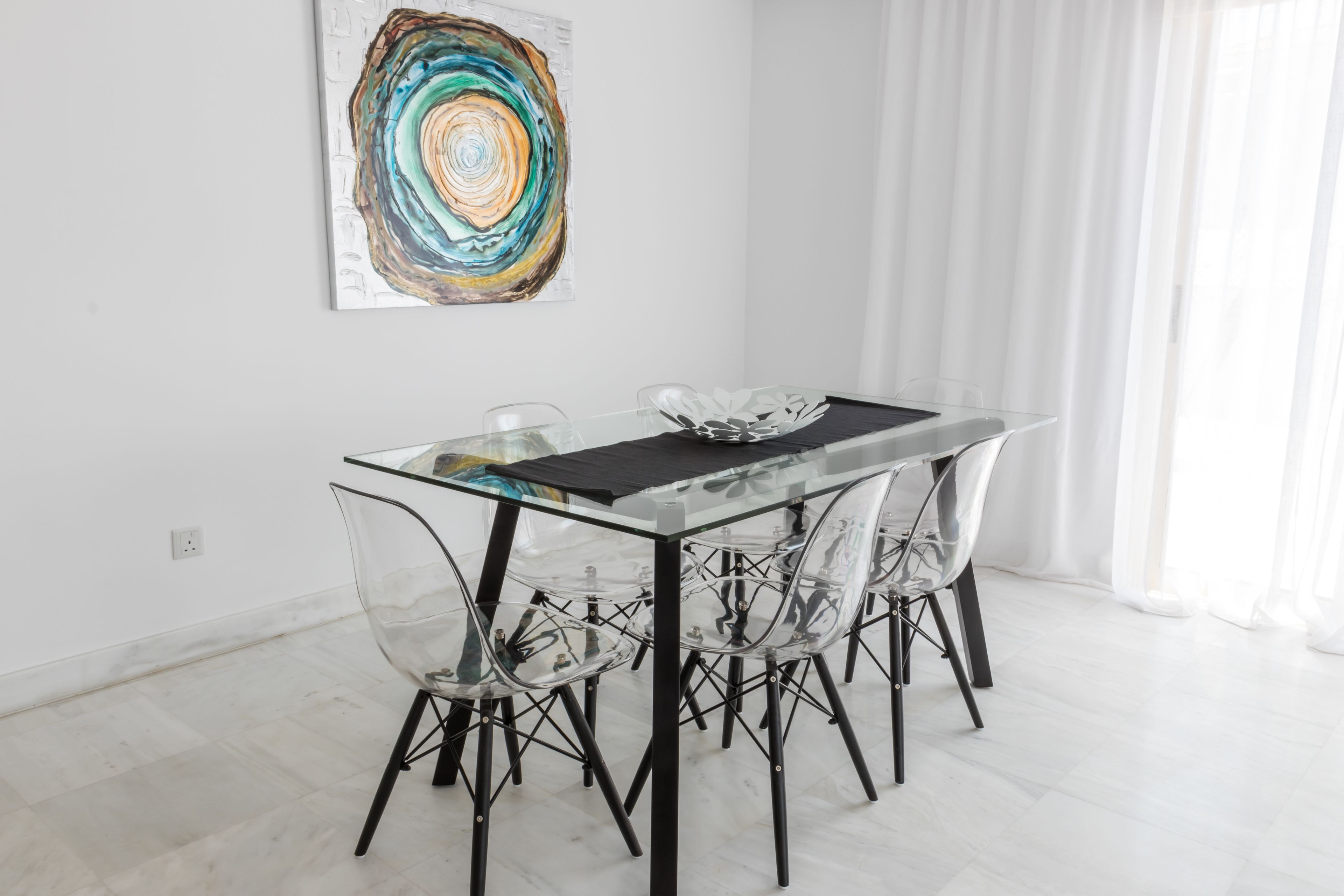 Uma área de refeições moderna com mesa de vidro e cadeiras transparentes, um sousbato preto e uma grande pintura abstrata na parede.
