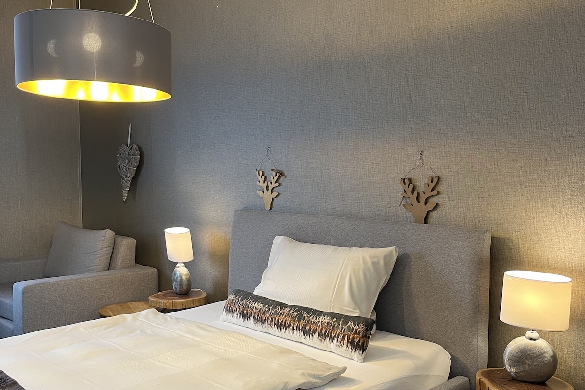 Quarto moderno com cama grande, duas mesas de cabeceira com luminárias, um sofá cinza estofado e um aplique na parede.