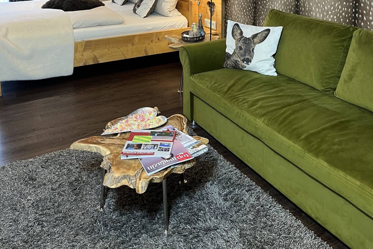 Quarto de hotel com uma cama, um sofá verde, uma mesa de centro de madeira com revistas e uma parede estampada.