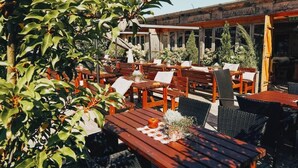 Um pátio externo com mesas e cadeiras de madeira, cercado por vegetação e uma fachada de construção rústica.