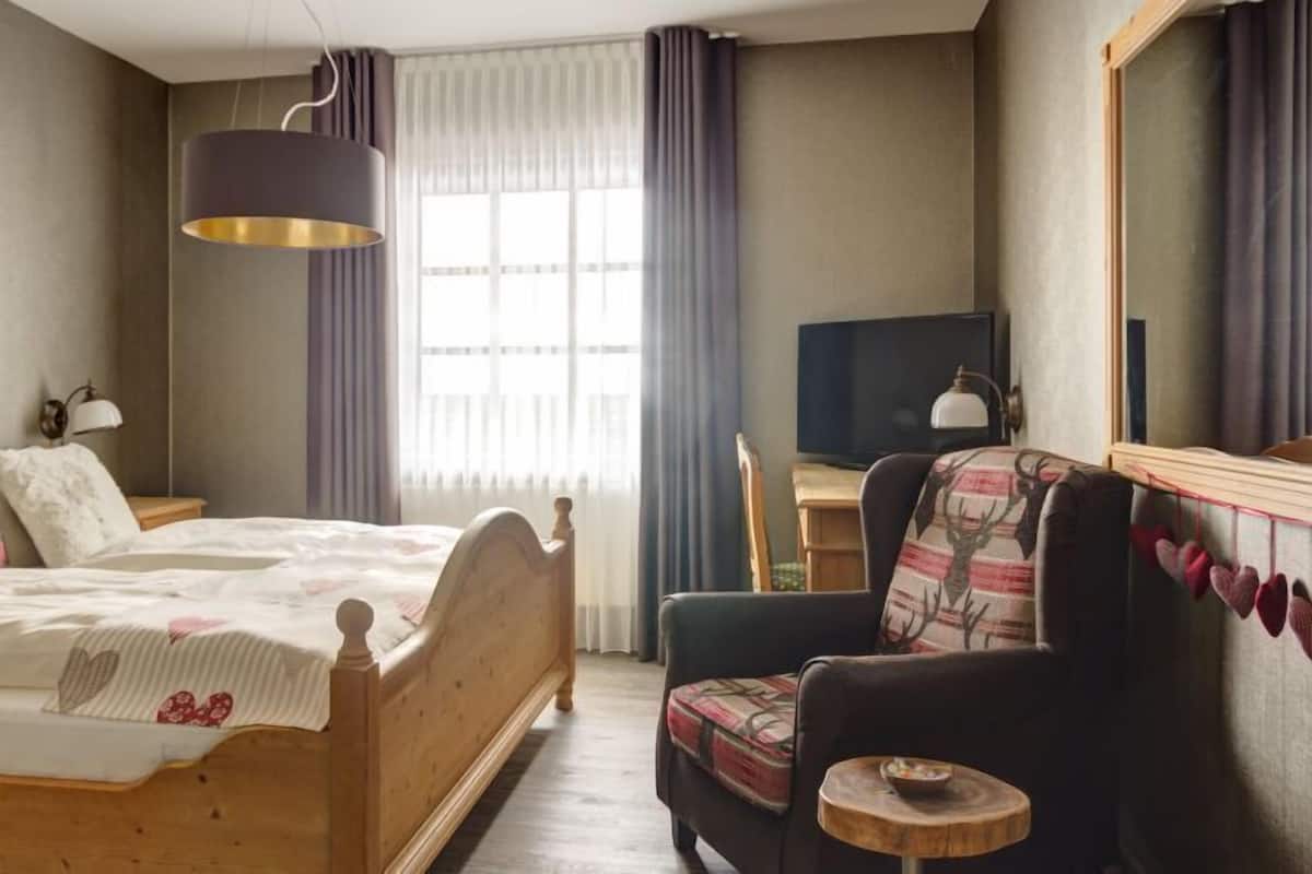 Quarto com cama de madeira, escrivaninha com cadeira, televisão e uma grande janela com cortinas.