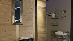 Sala de sauna com paredes de madeira, uma bacia preta grande, um espelho e uma prateleira de vime com toalhas.