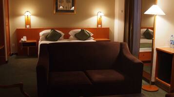 Quarto de hotel com sofá, mesa pequena, mesa de cabeceira, abajur e cama com almofadas.