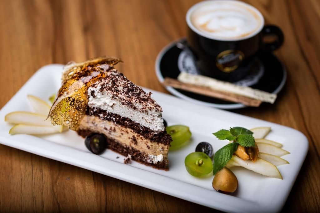 Uma fatia de bolo em camadas com chantilly em um prato branco, acompanhada de uma xícara de café e algumas frutas sobre uma mesa de madeira.