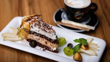 Uma fatia de bolo em camadas com chantilly em um prato branco, acompanhada de uma xícara de café e algumas frutas sobre uma mesa de madeira.