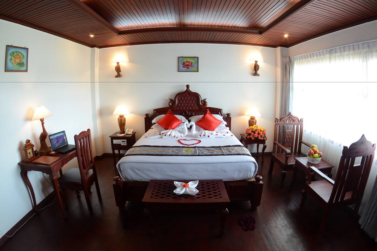 Quarto de hotel com uma cama grande, móveis de madeira, uma escrivaninha com um laptop e uma janela com cortinas.