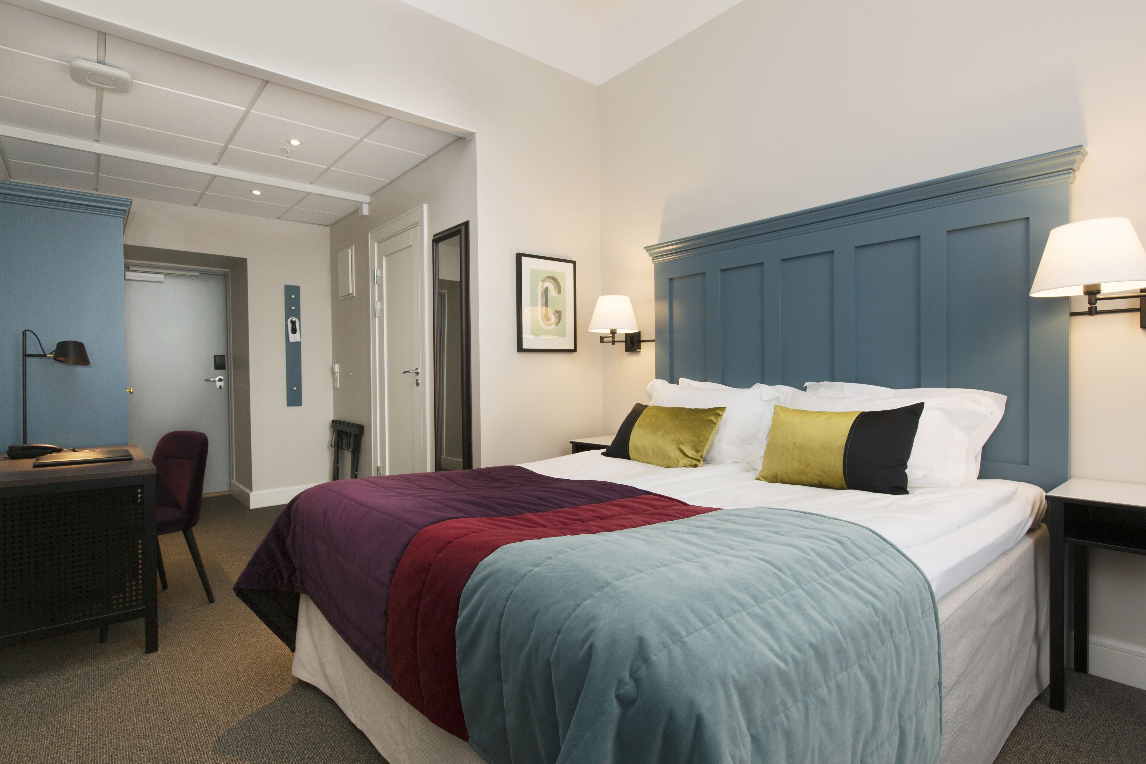 Quarto de hotel com cama, escrivaninha, cadeira e duas luminárias de cabeceira.