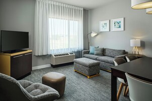 Quarto de hotel moderno com TV de tela plana, sofá, mesa de jantar e uma janela grande com cortinas leves.