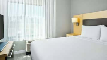 Quarto de hotel com cama, mesa de cabeceira, abajur, televisão e janela com cortinas finas.
