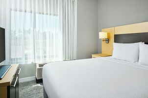 Quarto de hotel com cama, mesa de cabeceira, abajur, televisão e janela com cortinas finas.