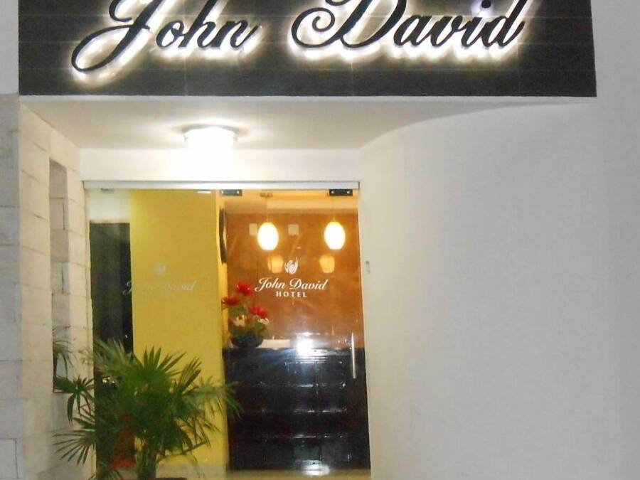 Entrada do Hotel John David com uma placa acima da porta e um vaso de planta em primeiro plano.