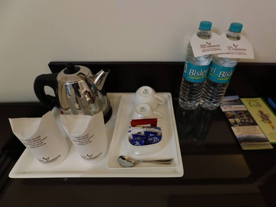 Uma bandeja com uma cafeteira, duas garrafas de água Bisleri, uma colher e um pequeno recipiente com embalagens, tudo sobre uma mesa.