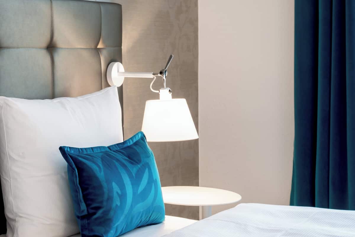 Cama bem arrumada com travesseiros brancos e azuis, mesa de cabeceira redonda e luminária fixada na parede.