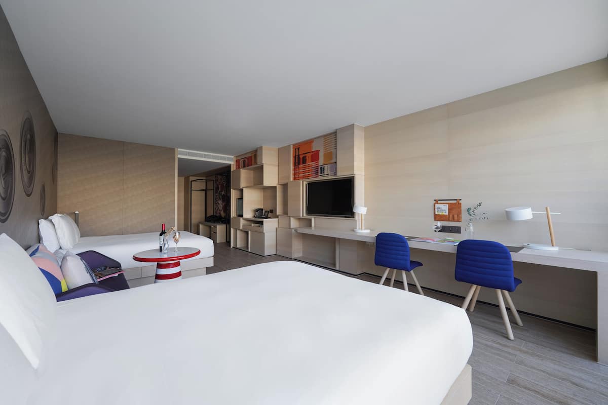 Quarto de hotel moderno com uma cama grande, uma escrivaninha com duas cadeiras, uma televisão e uma mesinha de cabeceira com um abajur.