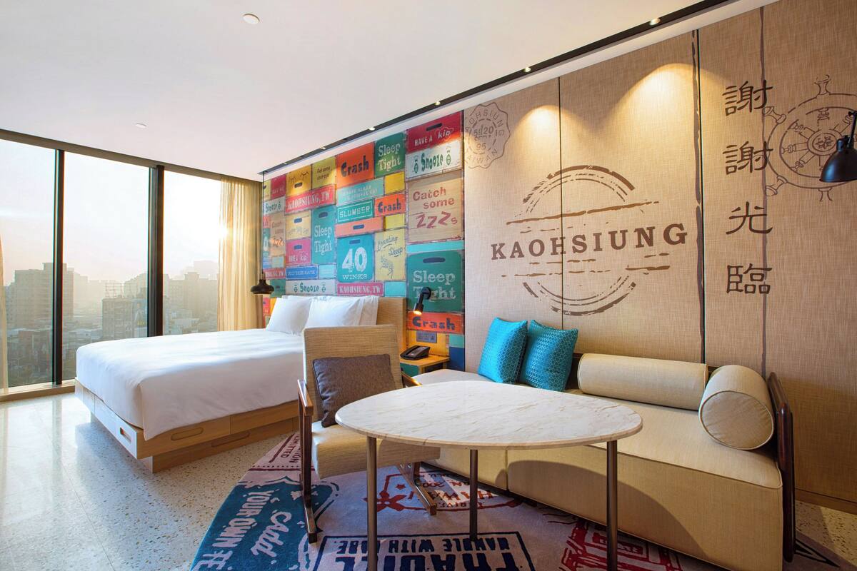 Quarto de hotel moderno com uma cama grande, um sofá e uma pequena mesa. O quarto oferece vista para a cidade através de amplas janelas e uma parede decorada com etiquetas de bagagem coloridas.