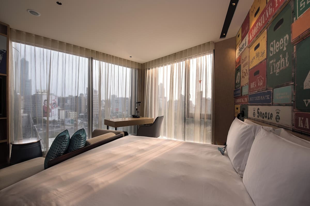 Quarto de hotel com uma cama grande, uma mesa de trabalho e vista para a cidade através de cortinas translúcidas.