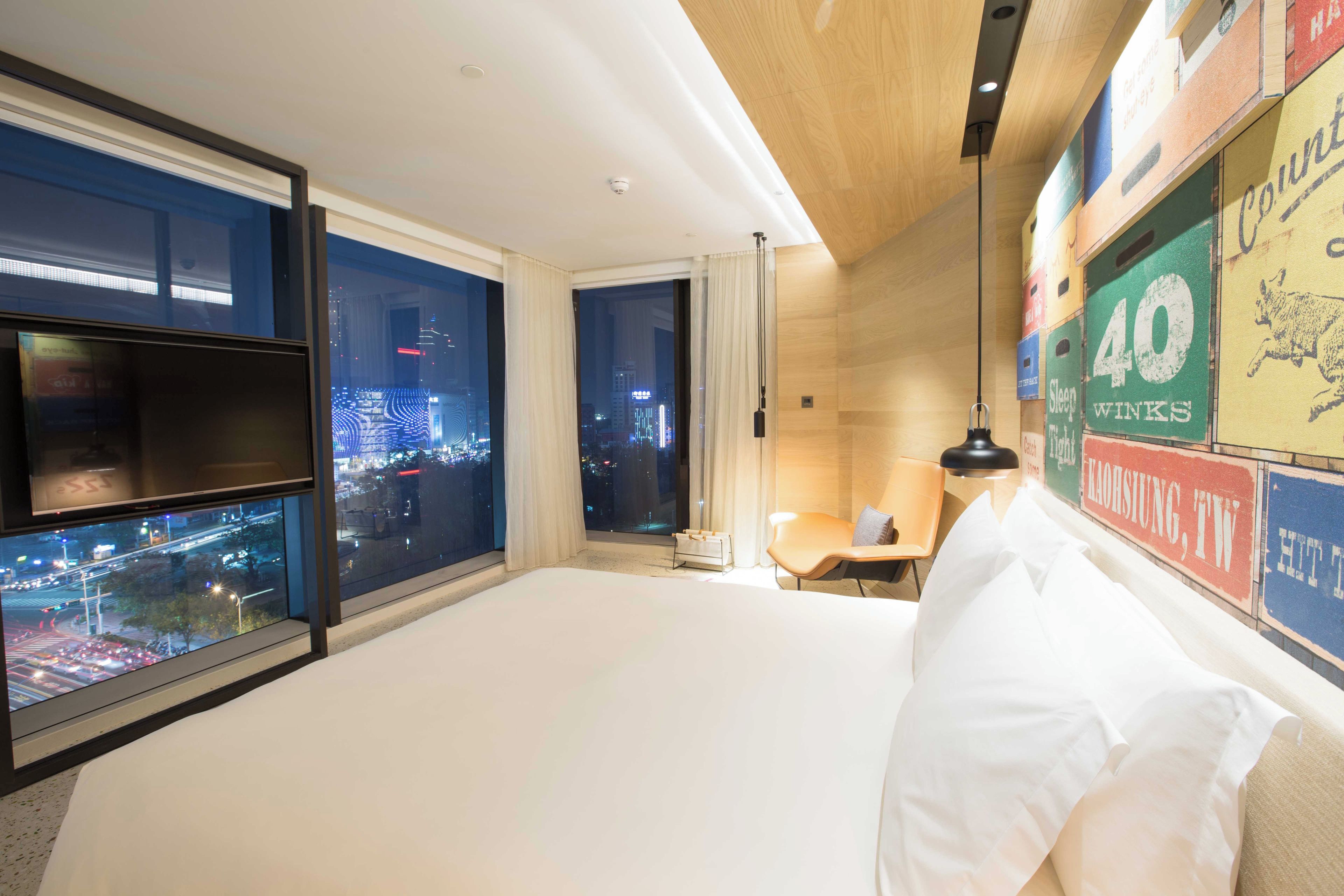 Quarto de hotel com uma cama grande, vista para a cidade pela janela, televisão fixada na parede e mesa de cabeceira.