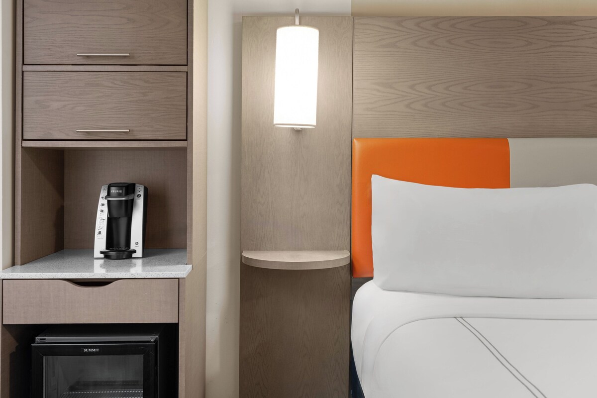 Quarto de hotel com cama, minibar, máquina de café e luminária fixada na parede.