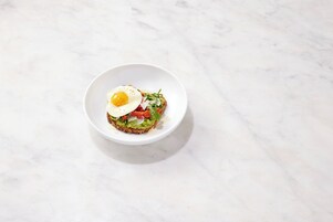 Uma tigela branca com um sanduíche de café da manhã, que leva ovo, abacate, tomate e folhas verdes sobre uma fatia de pão, sobre uma superfície de mármore.