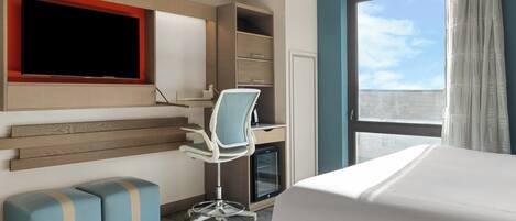Quarto de hotel com uma cama, uma secretária com cadeira, uma televisão e uma janela com cortinas.