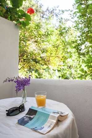 Uma mesa com um copo de suco de laranja, um vaso com flores roxas, óculos de sol e uma revista sobre uma toalha de mesa branca.