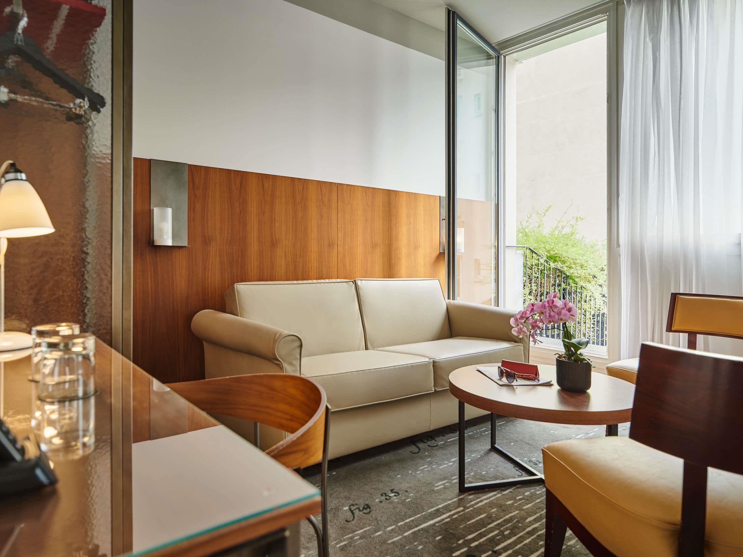 Quarto de hotel com sofá, mesa redonda, cadeira, luminária, pequena planta em vaso e grande janela com cortinas.