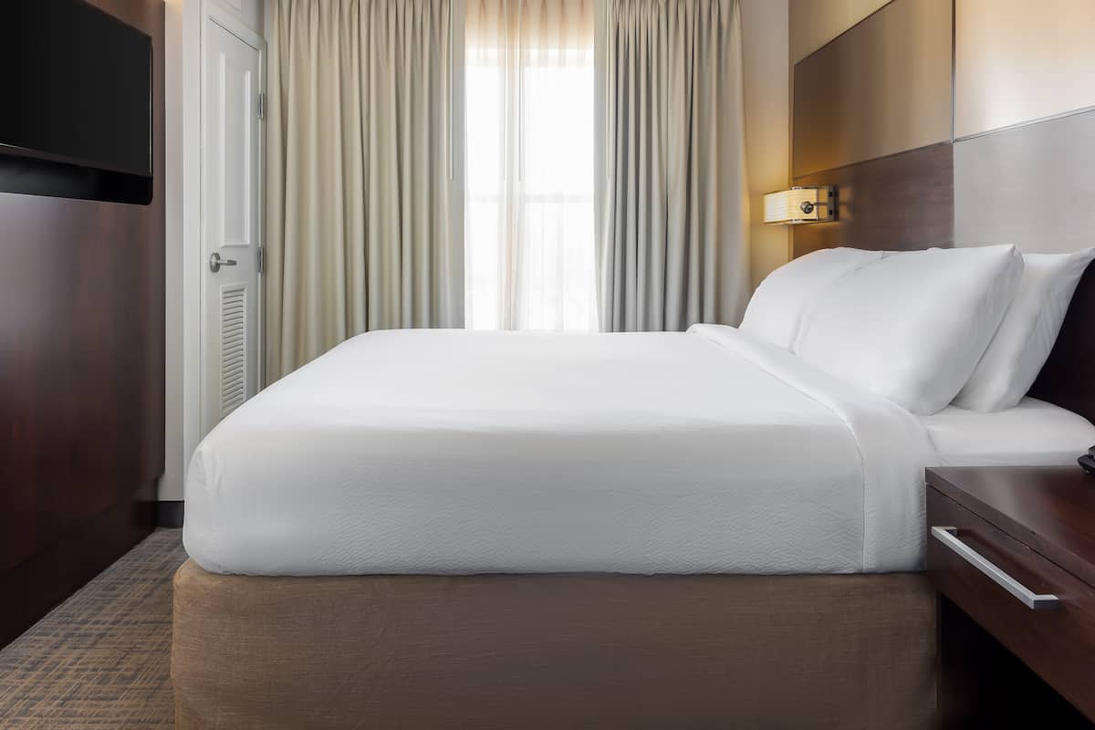Quarto de hotel com uma cama grande, televisão de tela plana na parede, mesa de cabeceira e janela com cortinas.