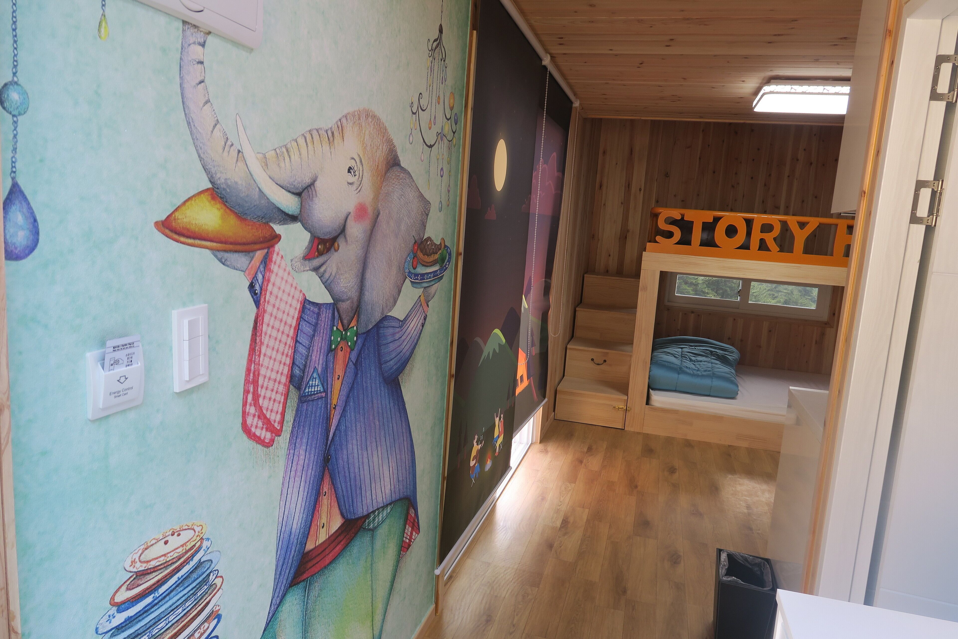 Quarto com um mural de elefante, beliches de madeira com a inscrição 'STORY' e corredor com piso de madeira.