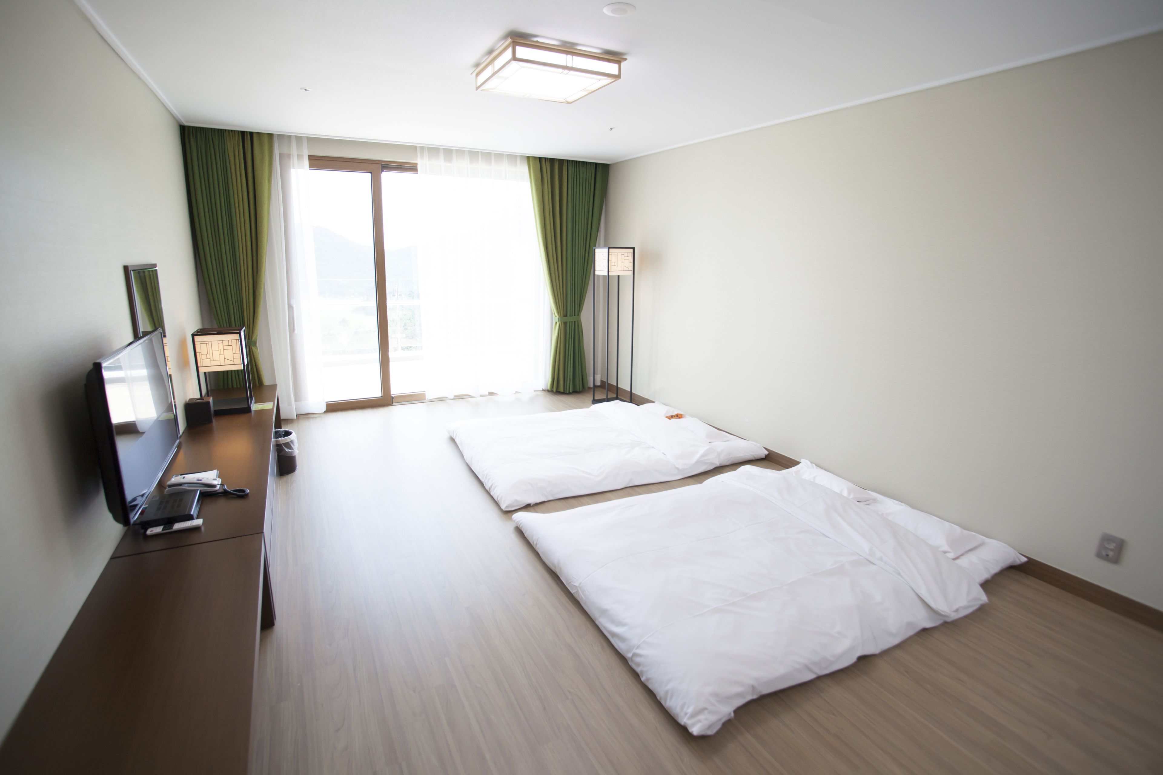 Quarto de hotel com uma cama grande, uma escrivaninha com um laptop, uma luminária e vista para o exterior.