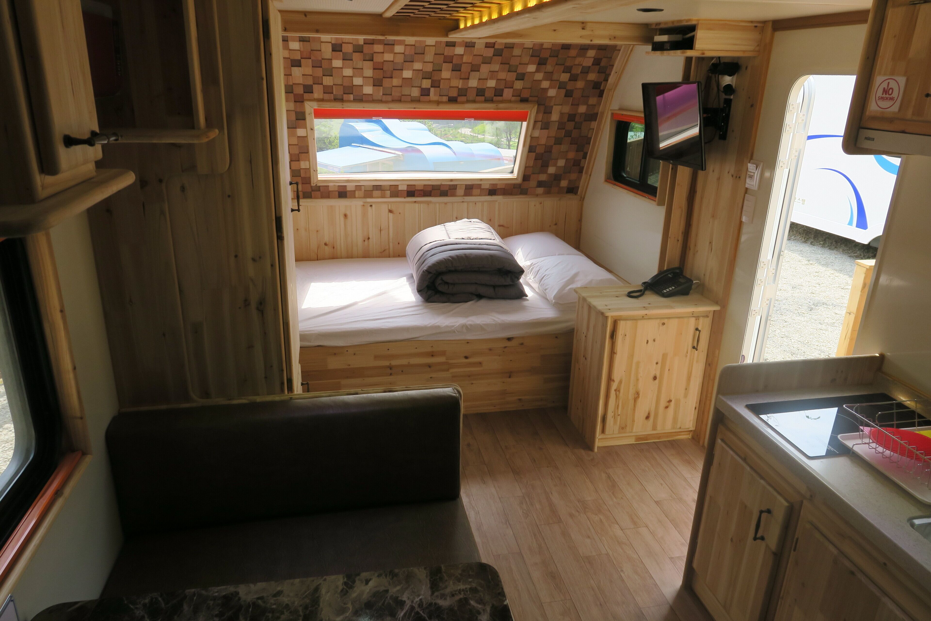 Interior compacto de uma motorhome, com cama, pequena cozinha, área de estar e janela com vista para um parquinho.