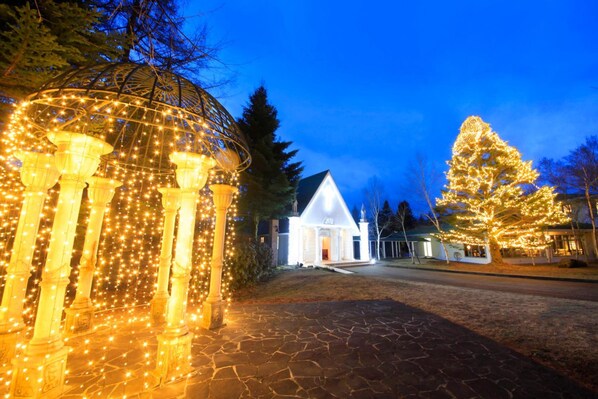 Um gazebo com luzes de corda, um edifício branco com telhado pontudo e uma árvore de Natal decorada.