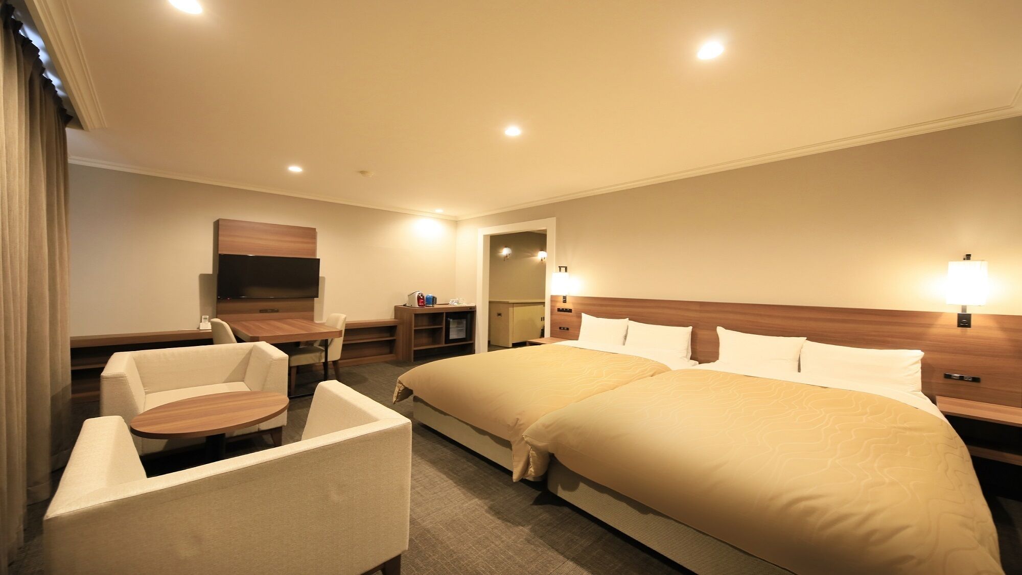 Quarto de hotel com duas camas, uma escrivaninha, uma cadeira, uma televisão e casa de banho.