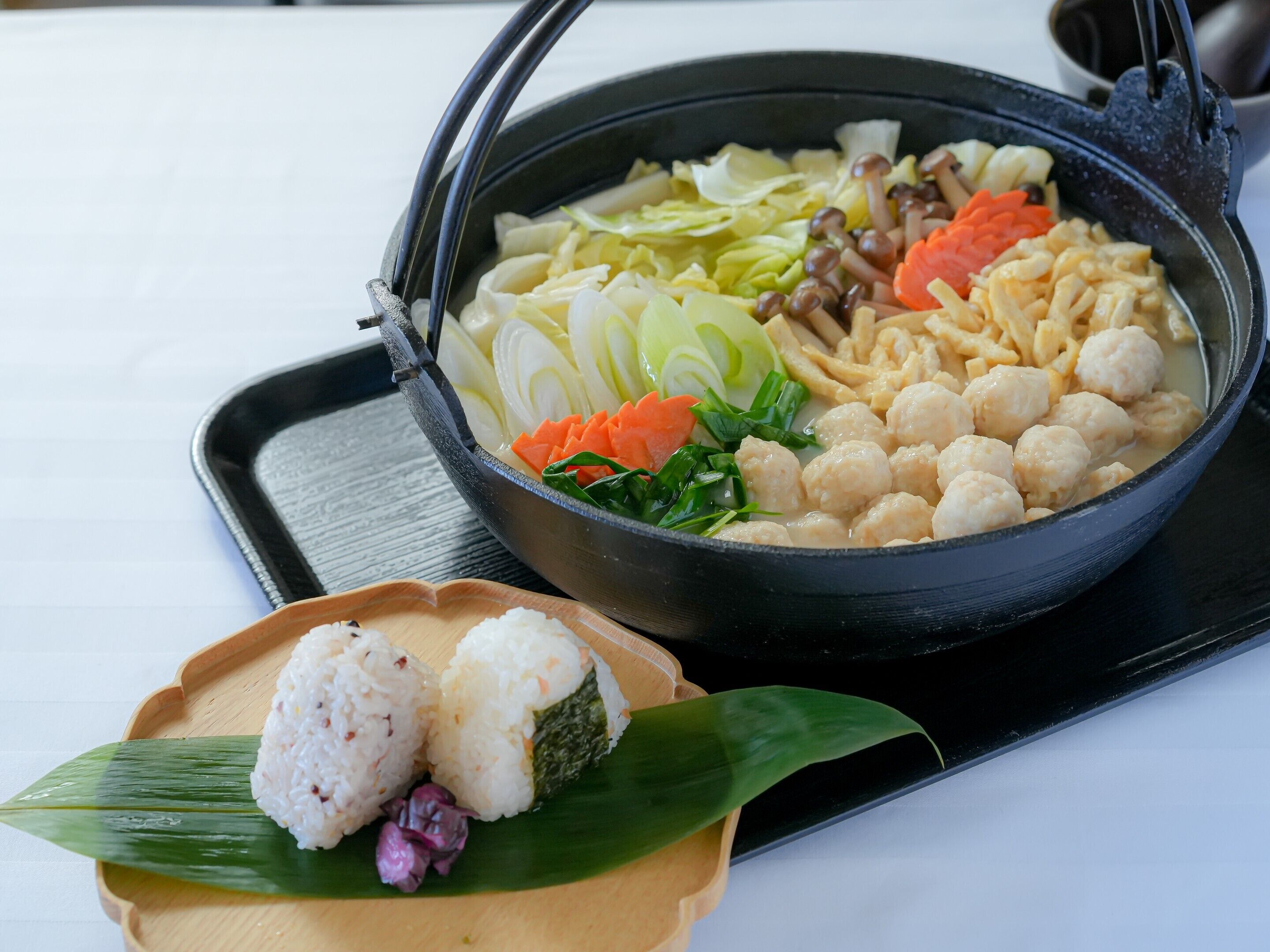 Uma tigela de sopa com diversos vegetais e dumplings, acompanhada de arroz e um pequeno prato de sushi.