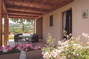 Um pátio externo com pérgola de madeira, uma mesa com cadeiras e vasos com flores cor-de-rosa.