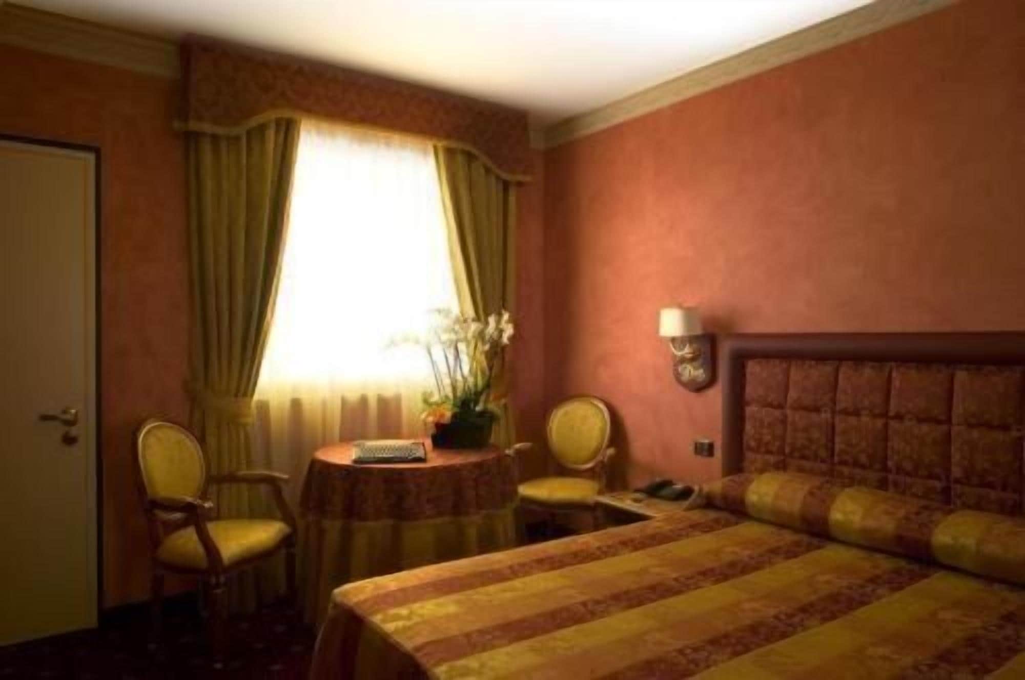 Quarto de hotel com uma cama grande, uma mesa redonda pequena com cadeiras, uma janela com cortinas e uma luminária na parede.