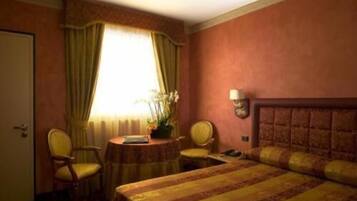 Quarto de hotel com uma cama grande, uma mesa redonda pequena com cadeiras, uma janela com cortinas e uma luminária na parede.