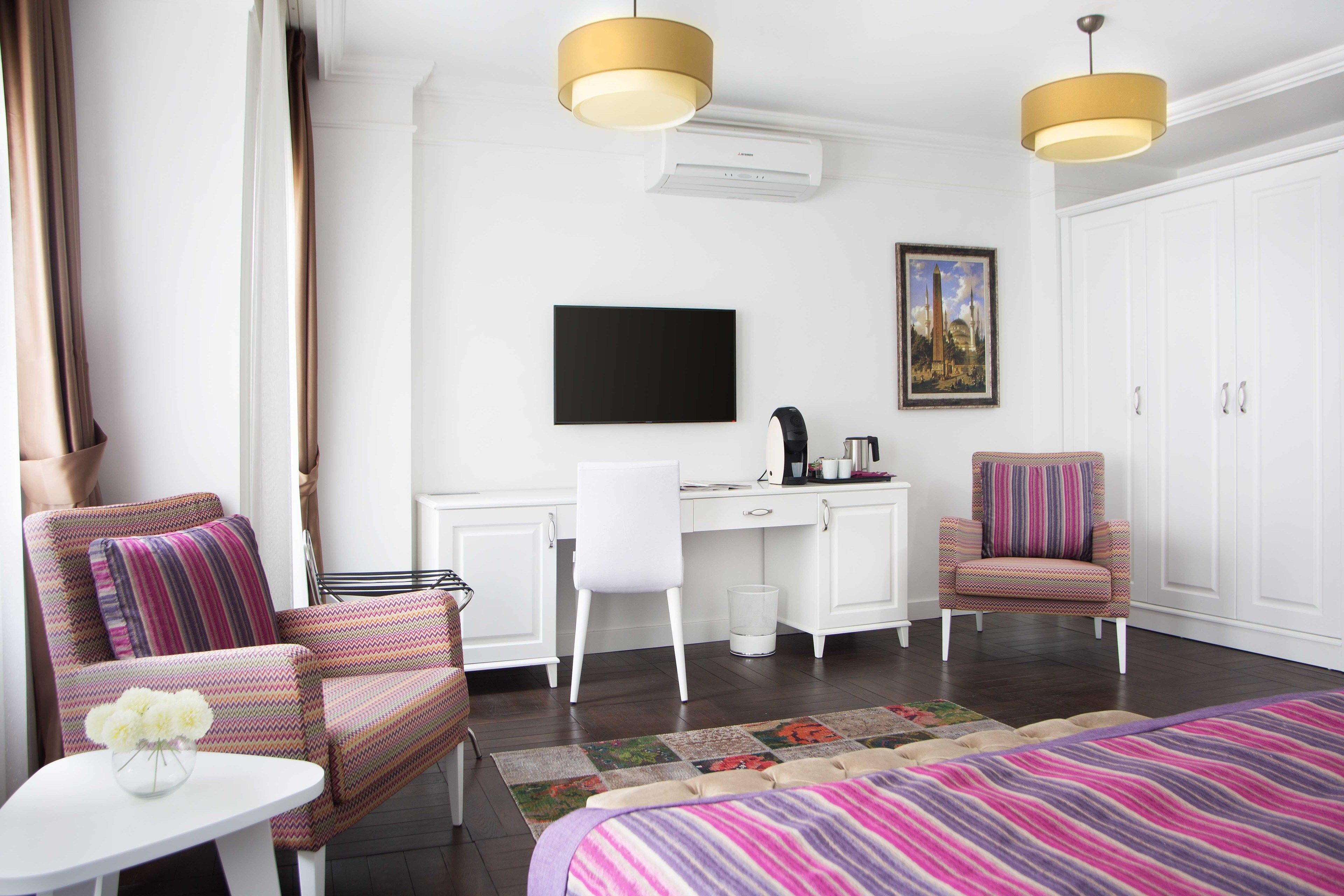 Quarto de hotel com cama, duas poltronas, escrivaninha, televisão e um quadro na parede.