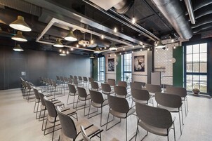 Sala de conferências com fileiras de cadeiras, amplas janelas e iluminação industrial.