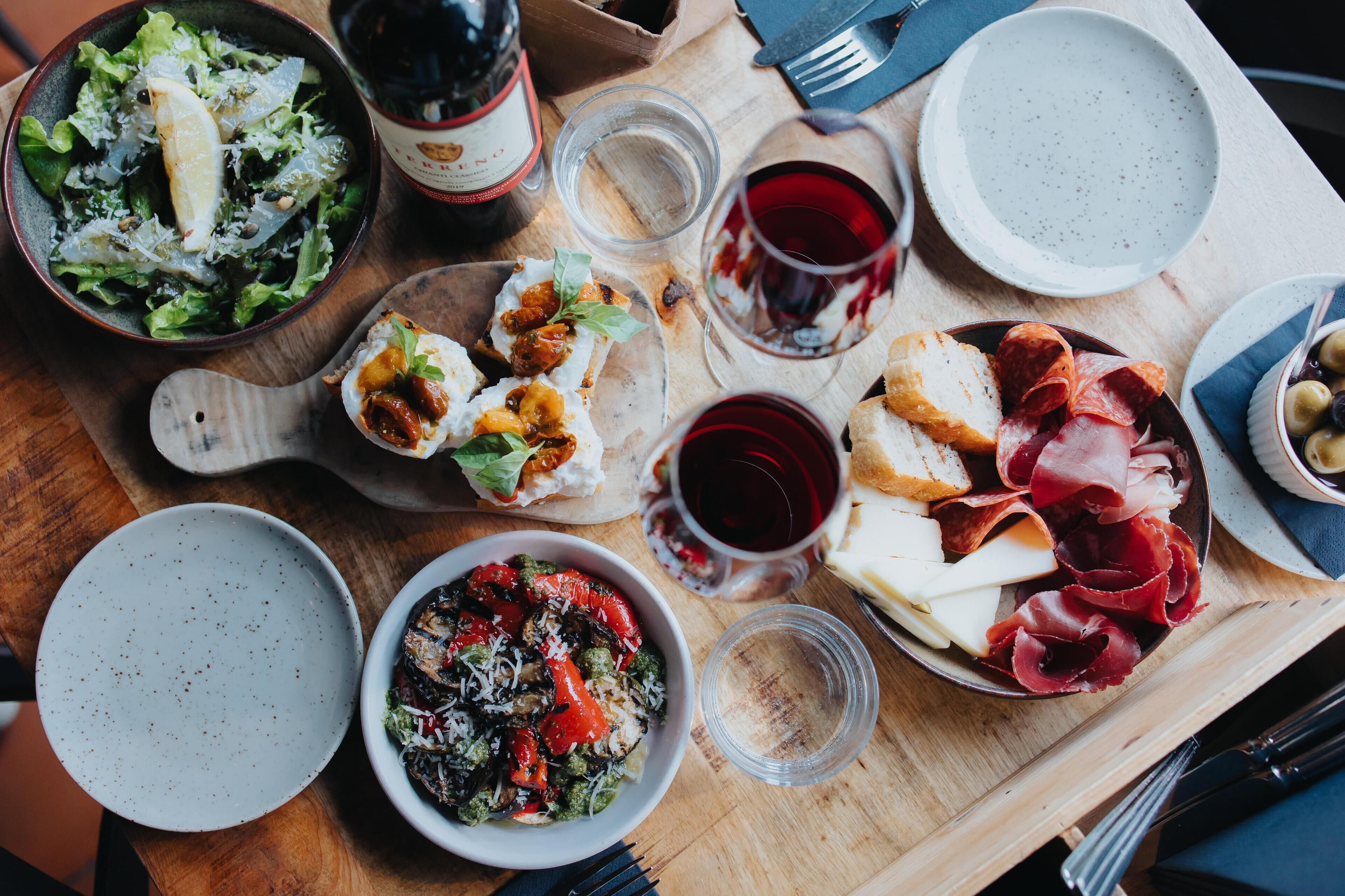Uma mesa de madeira com diversos alimentos e bebidas, incluindo uma saladeira, um prato de embutidos e copos de vinho tinto.