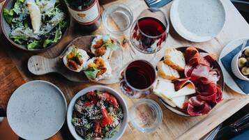 Uma mesa de madeira com diversos alimentos e bebidas, incluindo uma saladeira, um prato de embutidos e copos de vinho tinto.