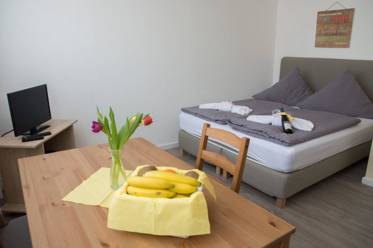Quarto de hotel bem arrumado, com uma cama, uma mesinha de cabeceira com controle remoto, uma pequena mesa com um cesto de bananas e flores, e uma parede com um pôster.