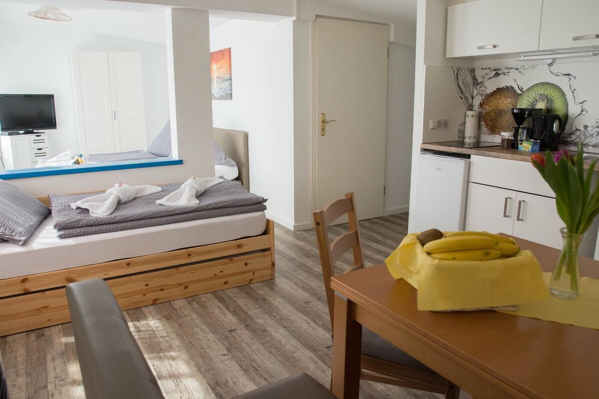 Quarto com cama de madeira, mesa de cabeceira, cadeira, pequena mesa com fruteira e área de cozinha com cafeteira e frutas.