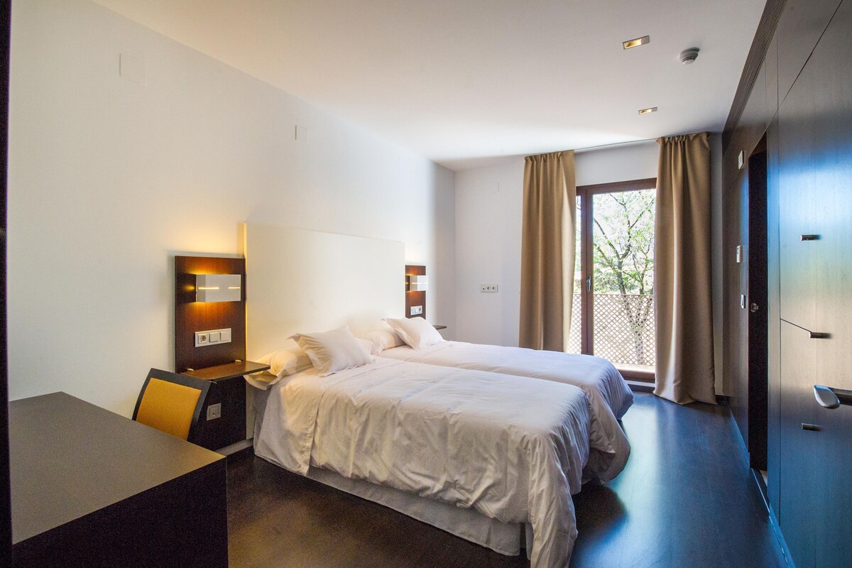 Quarto de hotel com duas camas, uma escrivaninha e uma janela grande com cortinas.