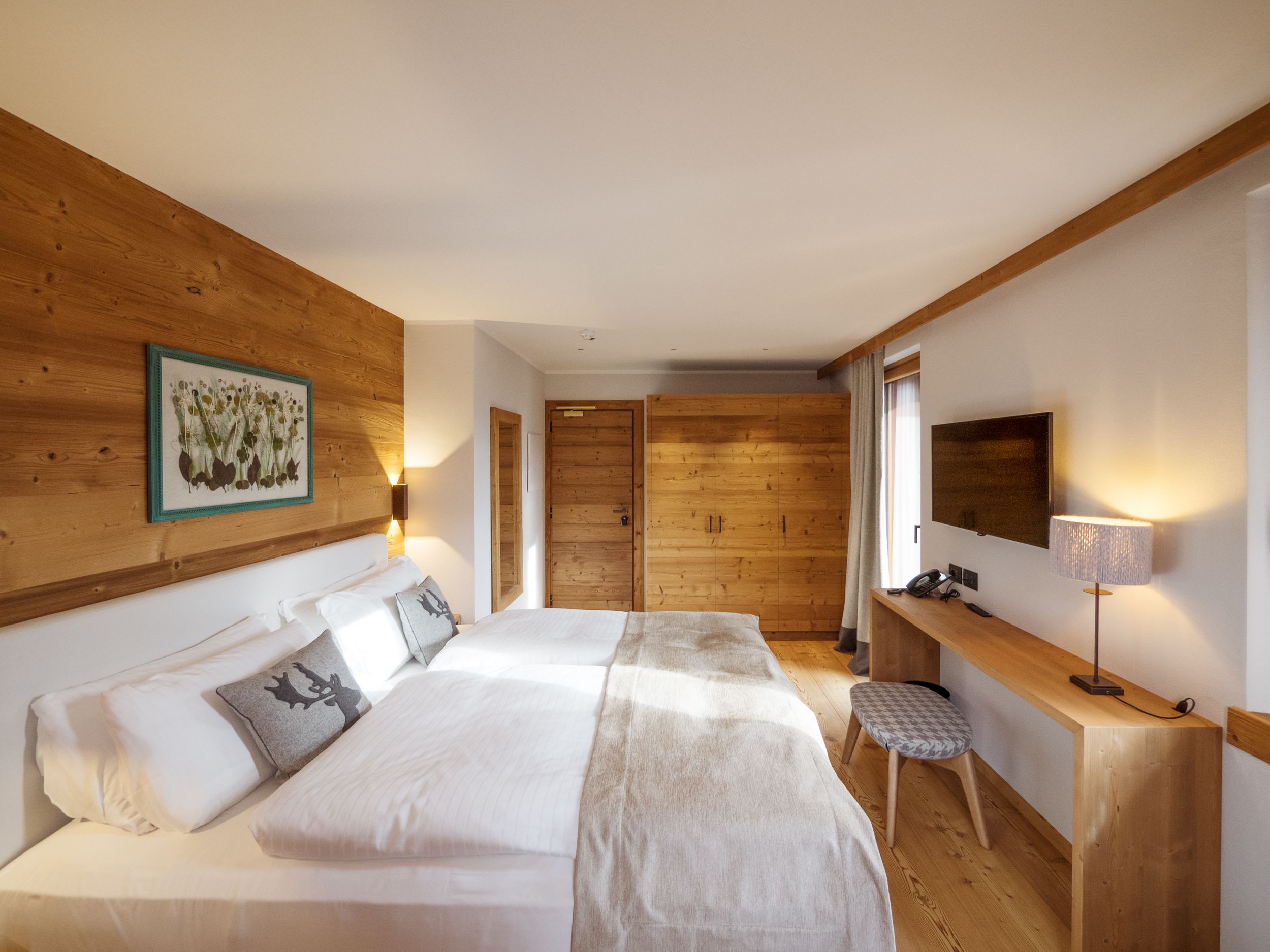 Quarto com cabeceira de madeira, cama com roupa de cama branca, mesa de cabeceira com abajur, escrivaninha com cadeira e um espelho grande na parede.