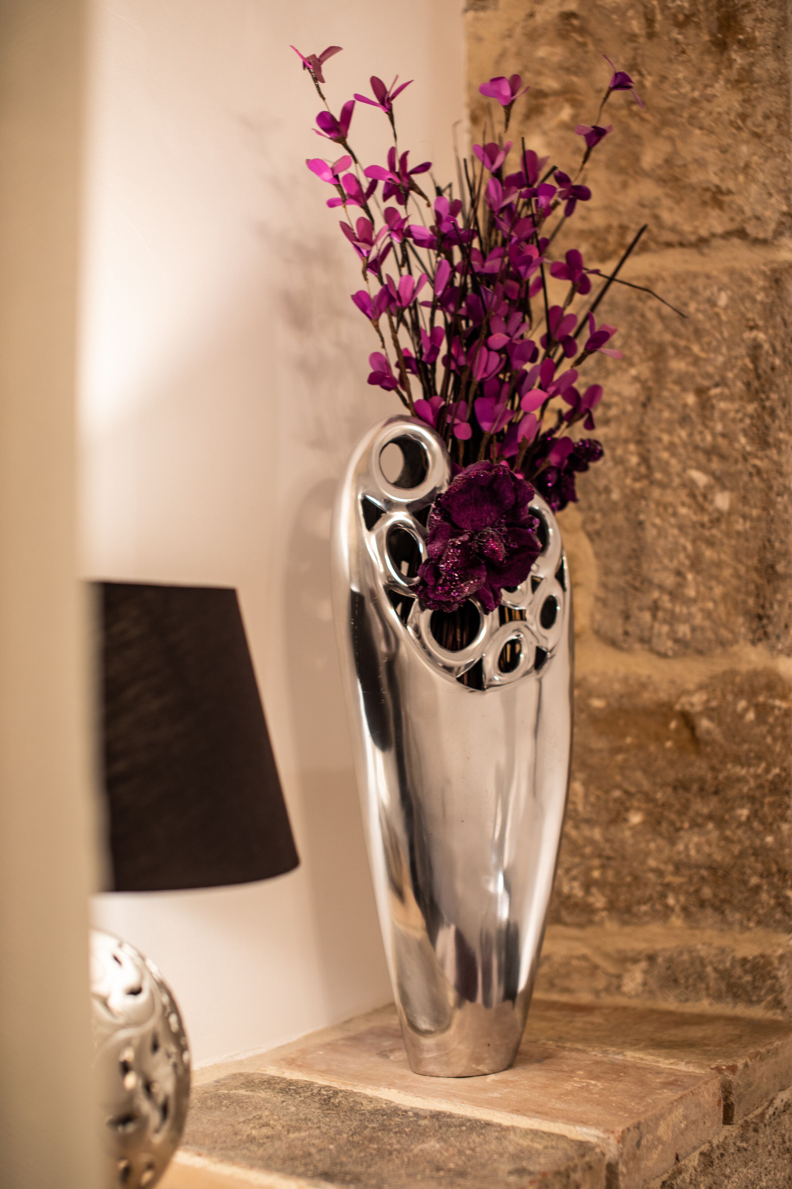 Um vaso metálico com design único, contendo flores roxas, apoiado em um parapeito de pedra contra uma parede.