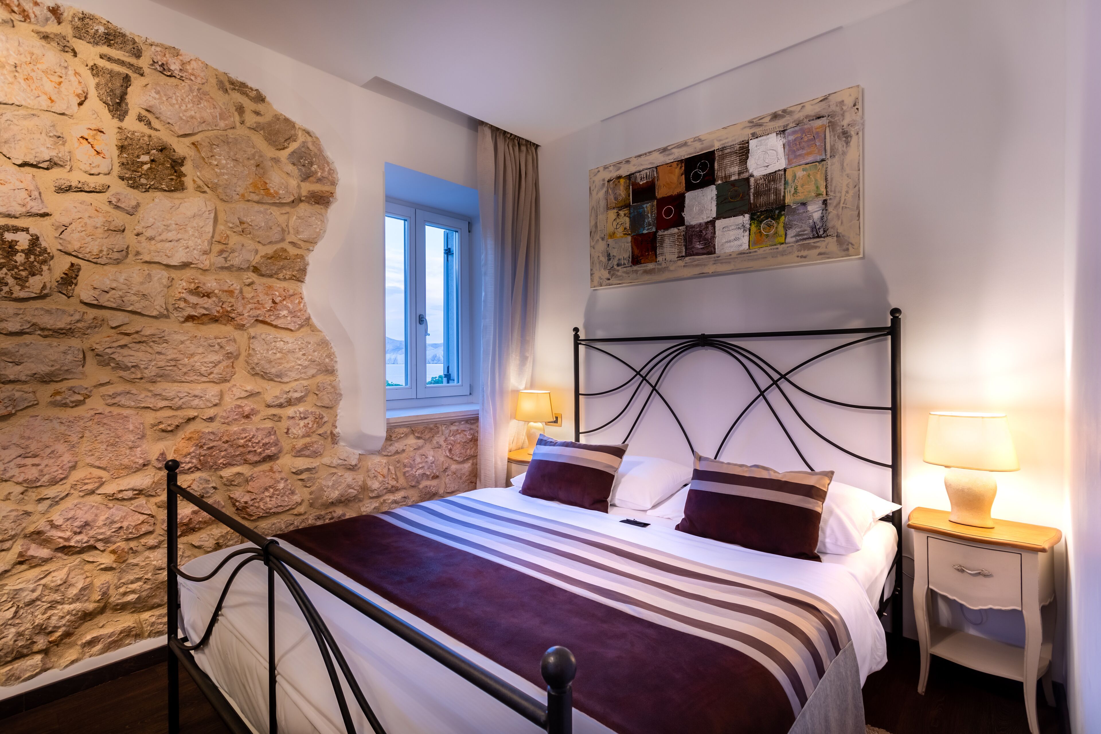 Quarto com parede de pedra, cama com roupa de cama listrada, cabeceira, luminárias de cabeceira e janela com cortinas.