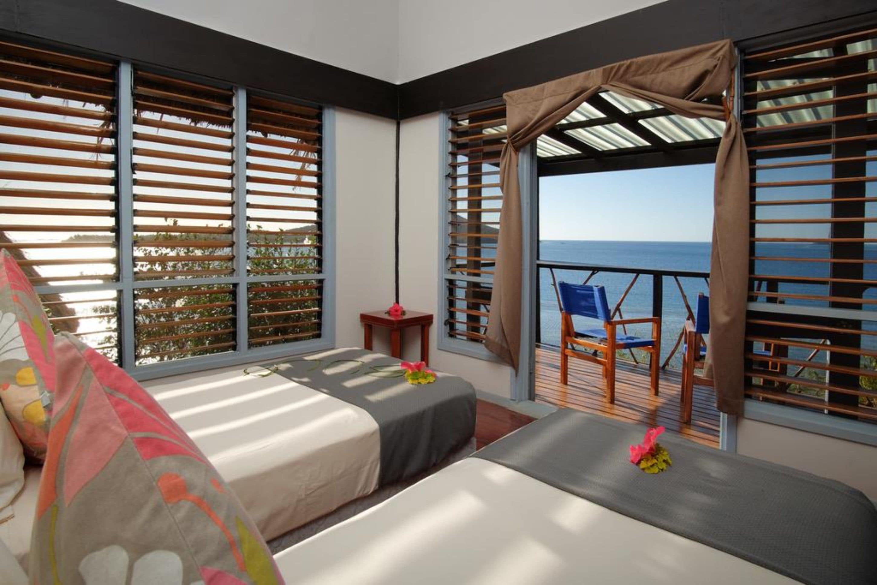 Quarto com duas camas, uma mesa pequena e uma cadeira, com vista para o mar.